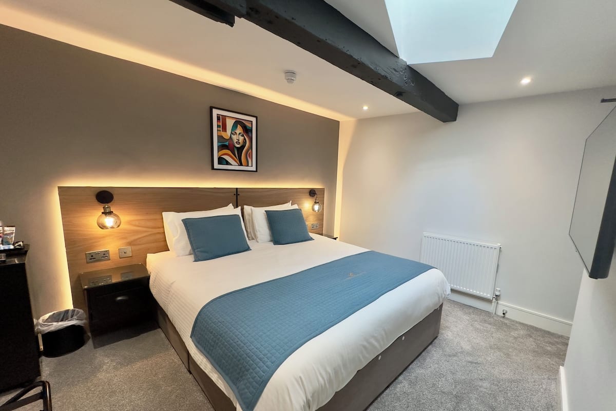 Quarto moderno com uma cama grande, cabeceira com luminárias fixadas na parede, ventilador de teto e claraboia.