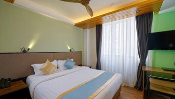 Quarto de hotel com cama, cabeceira de madeira, ventilador de teto, televisão e escrivaninha com cadeira.