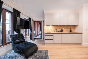 Um apartamento moderno com um sofá preto estofado, uma pequena mesa lateral, uma cozinha com armários brancos e bancada de madeira, e uma porta de vidro deslizante que dá para uma varanda.