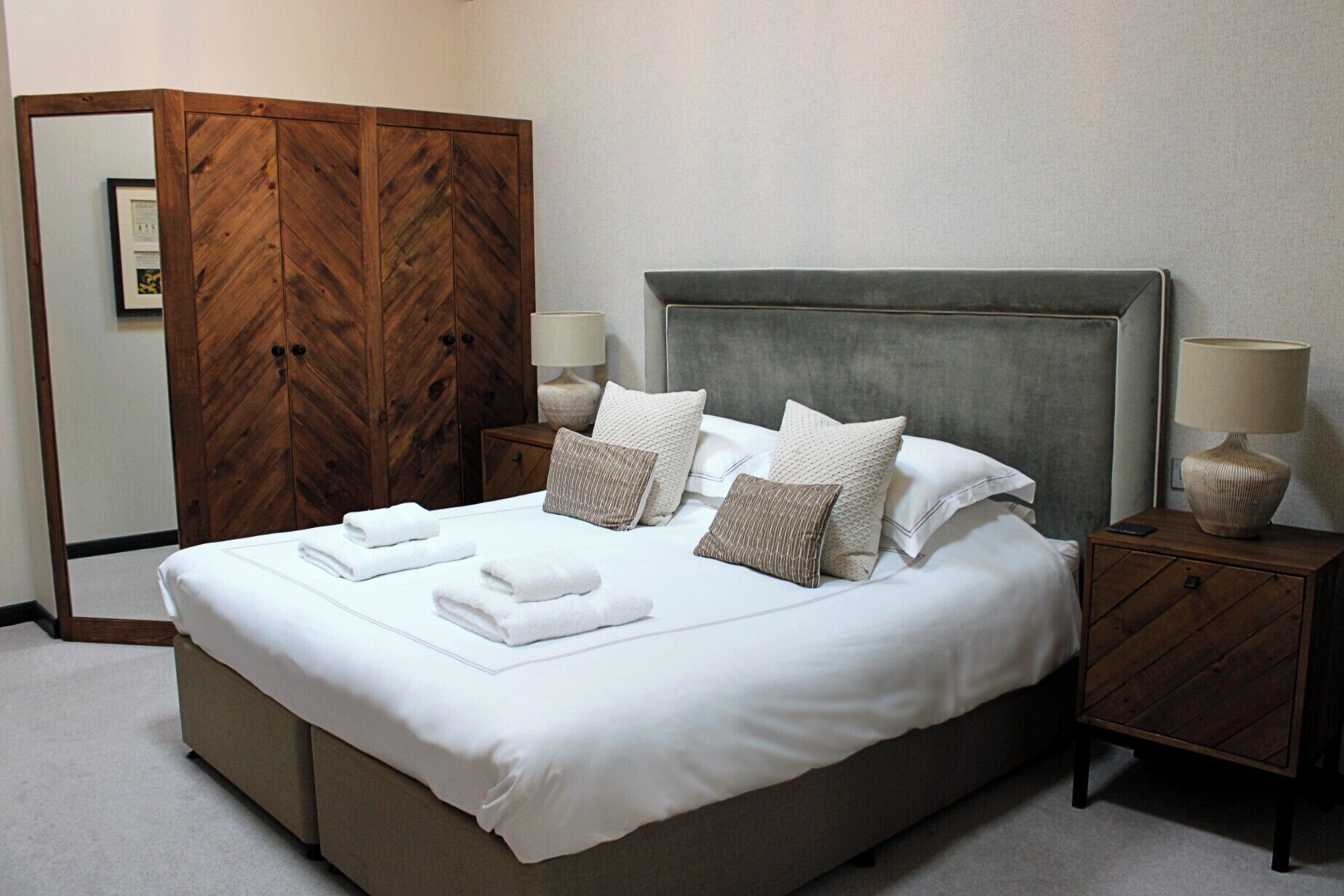 Cama bem arrumada com lençóis brancos e várias almofadas, guarda-roupa de madeira e duas mesas de cabeceira com abajures.