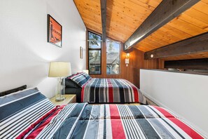 Quarto com duas camas, teto de madeira, janela e luminária fixada na parede.