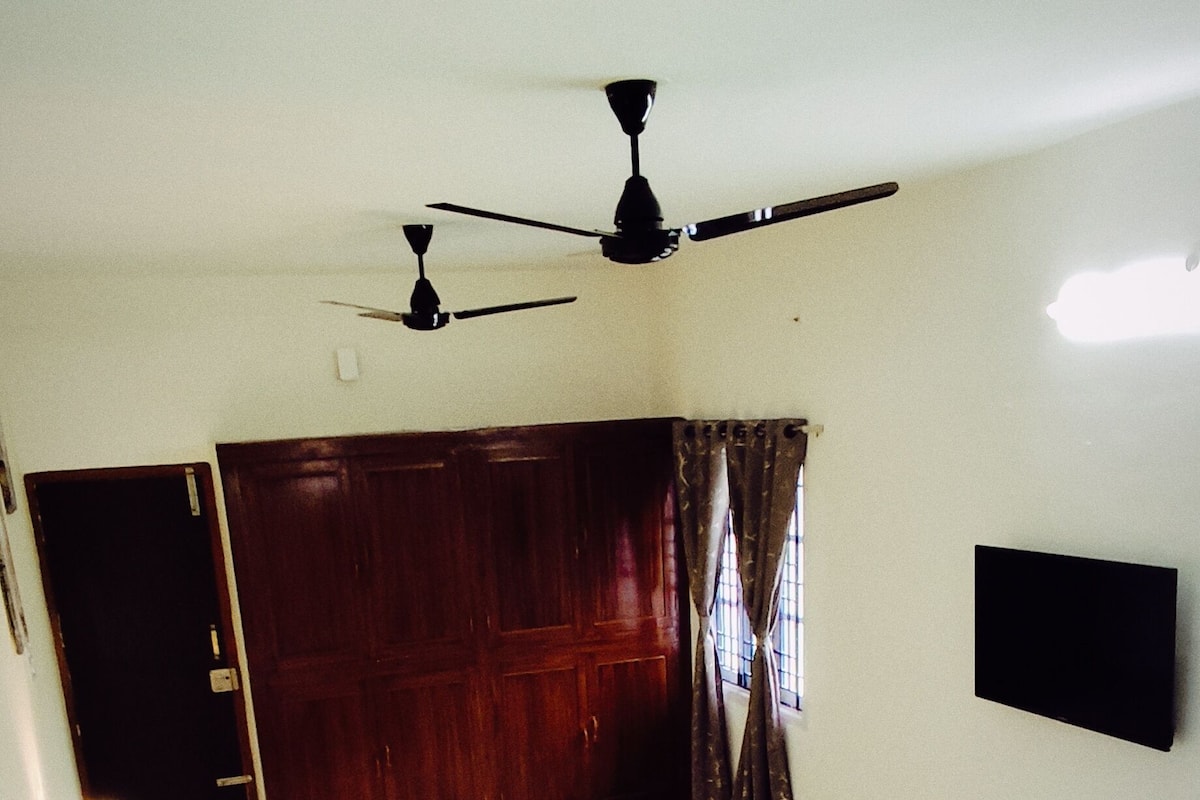 Uma cama de solteiro com cabeceira, mesa de cabeceira, cadeira, ventoinha no teto e televisão fixada na parede.