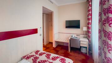 Quarto de hotel com cama, escrivaninha, cadeira, televisão e porta que dá para outro cômodo.