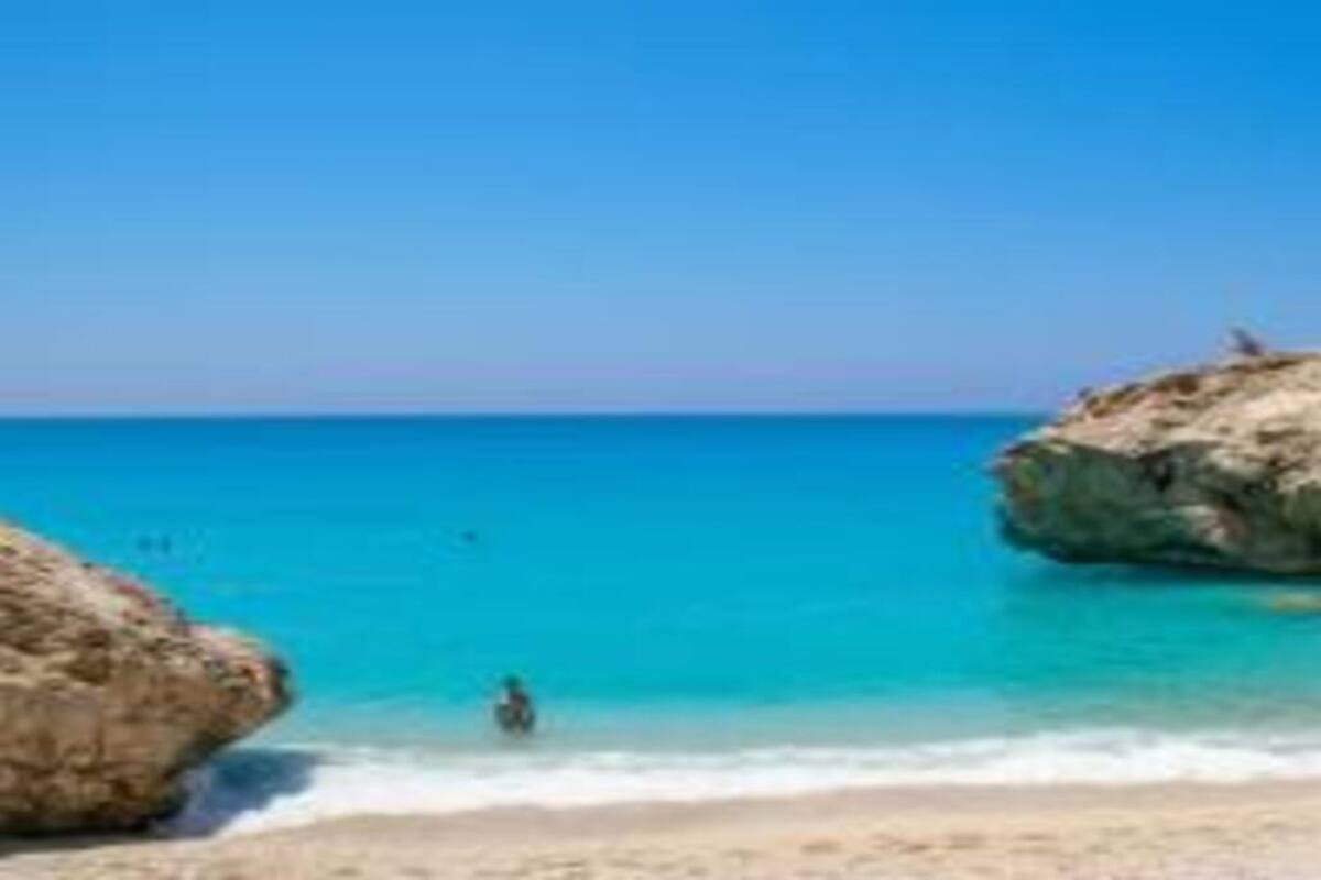 Uma praia com águas azul-claras, uma grande formação rochosa e uma pessoa na água.