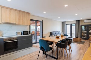 Cozinha moderna com eletrodomésticos embutidos, sala de jantar com mesa e cadeiras de madeira, e área de estar com sofá e cortinas.