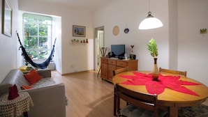 Uma área de refeições com uma mesa de madeira, cadeiras e um jogo americano vermelho. Há uma televisão em um armário de madeira e uma rede ao fundo.