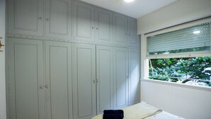 Quarto com guarda-roupa espaçoso, cama com colcha florida e janela com persianas.