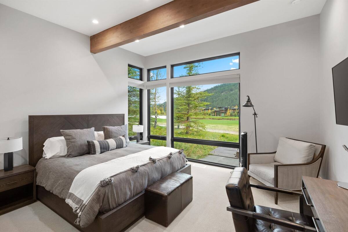 Quarto moderno com cama grande, escrivaninha e cadeira, com vista para uma paisagem montanhosa através de amplas janelas.