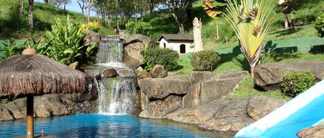 Piscina transparente com cascata, rodeada por pedras e vegetação.