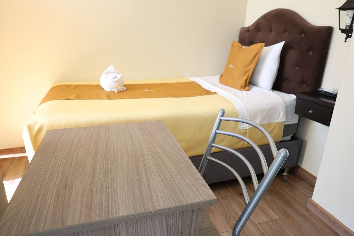 Cama bem arrumada com cabeceira marrom, manta amarela e travesseiro branco. Tem uma pequena mesa de cabeceira com luminária e gaveta. Uma cadeira de metal está posicionada em frente à cama.