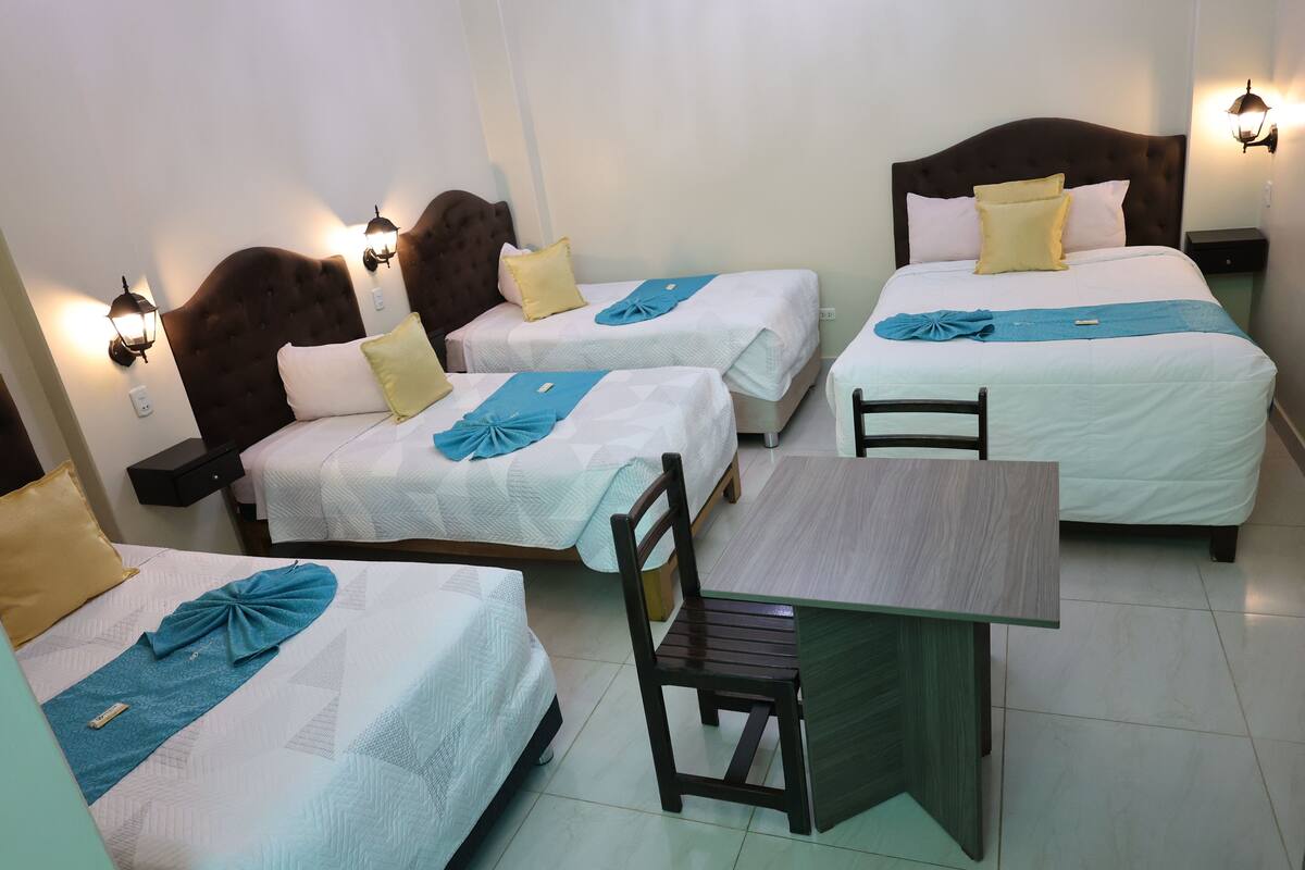 Quarto de hotel com três camas, cada uma com cabeceira e luminárias laterais. Há uma mesa de madeira com uma cadeira no centro.