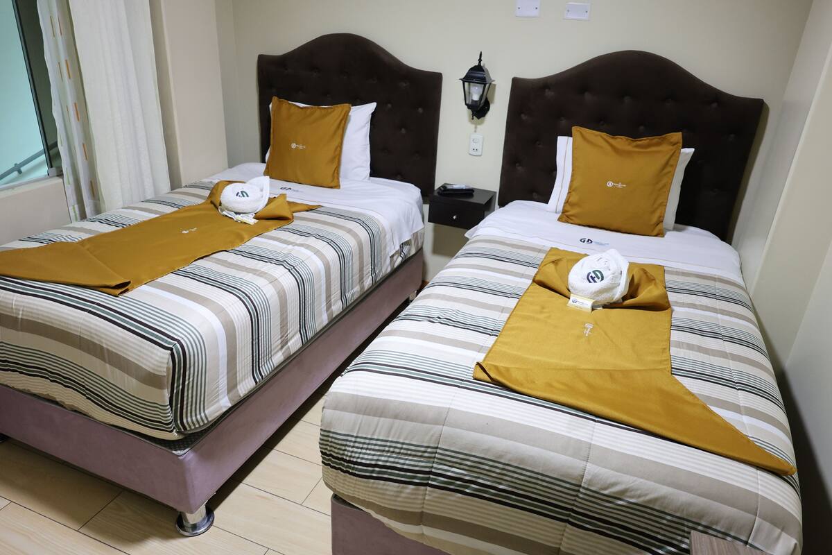 Duas camas com roupa de cama listrada e cabeceiras marrons, uma mesa de cabeceira com abajur e uma janela com cortinas.