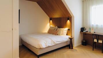 Quarto com teto inclinado, cama com roupa de cama branca, escrivaninha com cadeira e janela com cortinas.
