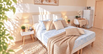 Quarto com cama, mesas de cabeceira, um criado-mudo, luminária, pequena mesa com uma planta e geladeira.