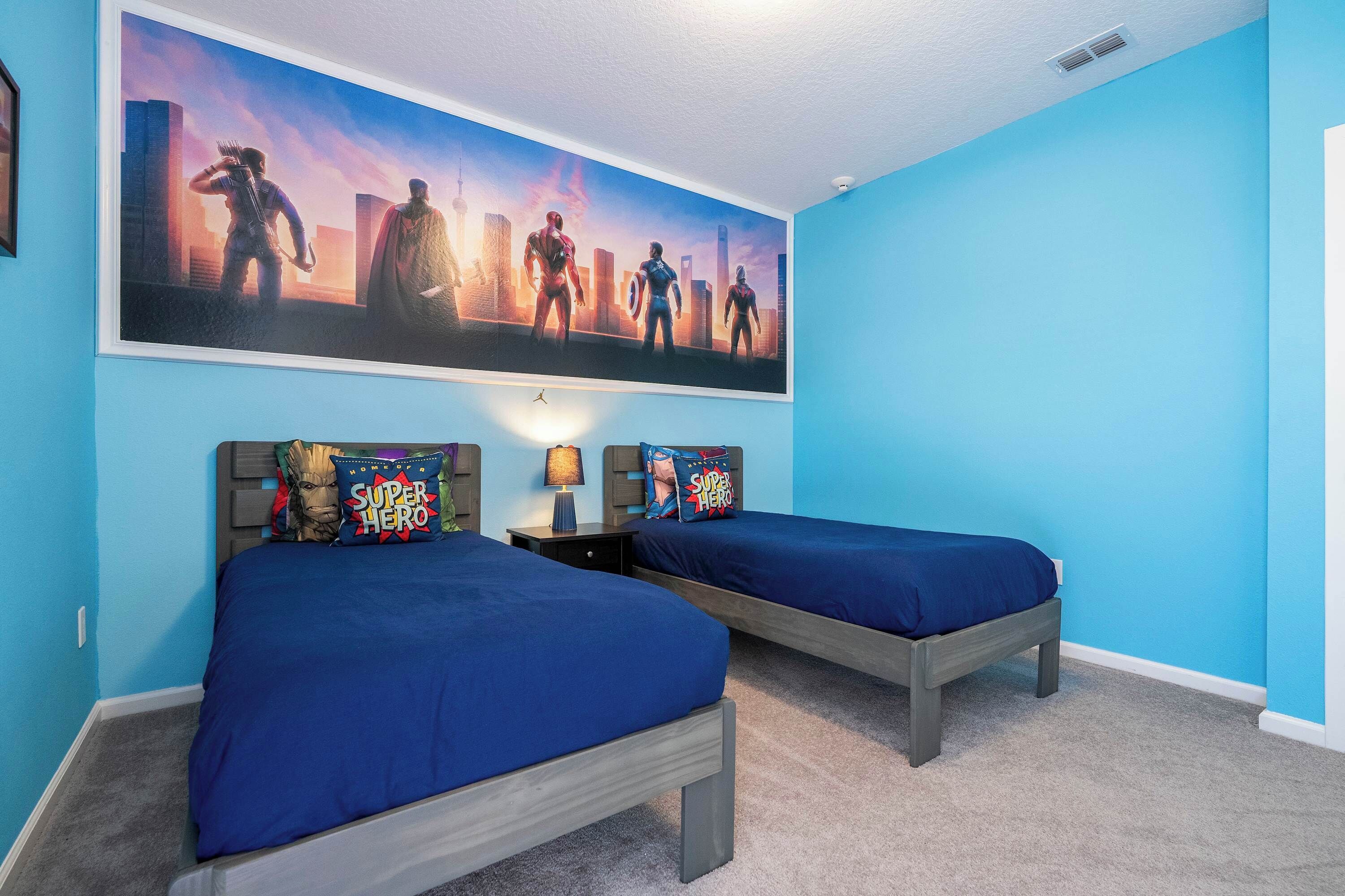 Quarto com duas camas, um pôster grande com tema de super-herói e uma mesa de cabeceira com abajur.