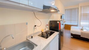 Cozinha moderna com pia de aço inoxidável, fogão a gás e geladeira. Há uma janela pequena com persianas e, no quarto adjacente, uma cama com colcha branca.
