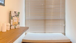 Quarto com mesa de cabeceira de madeira, colchão branco e janela com persianas.