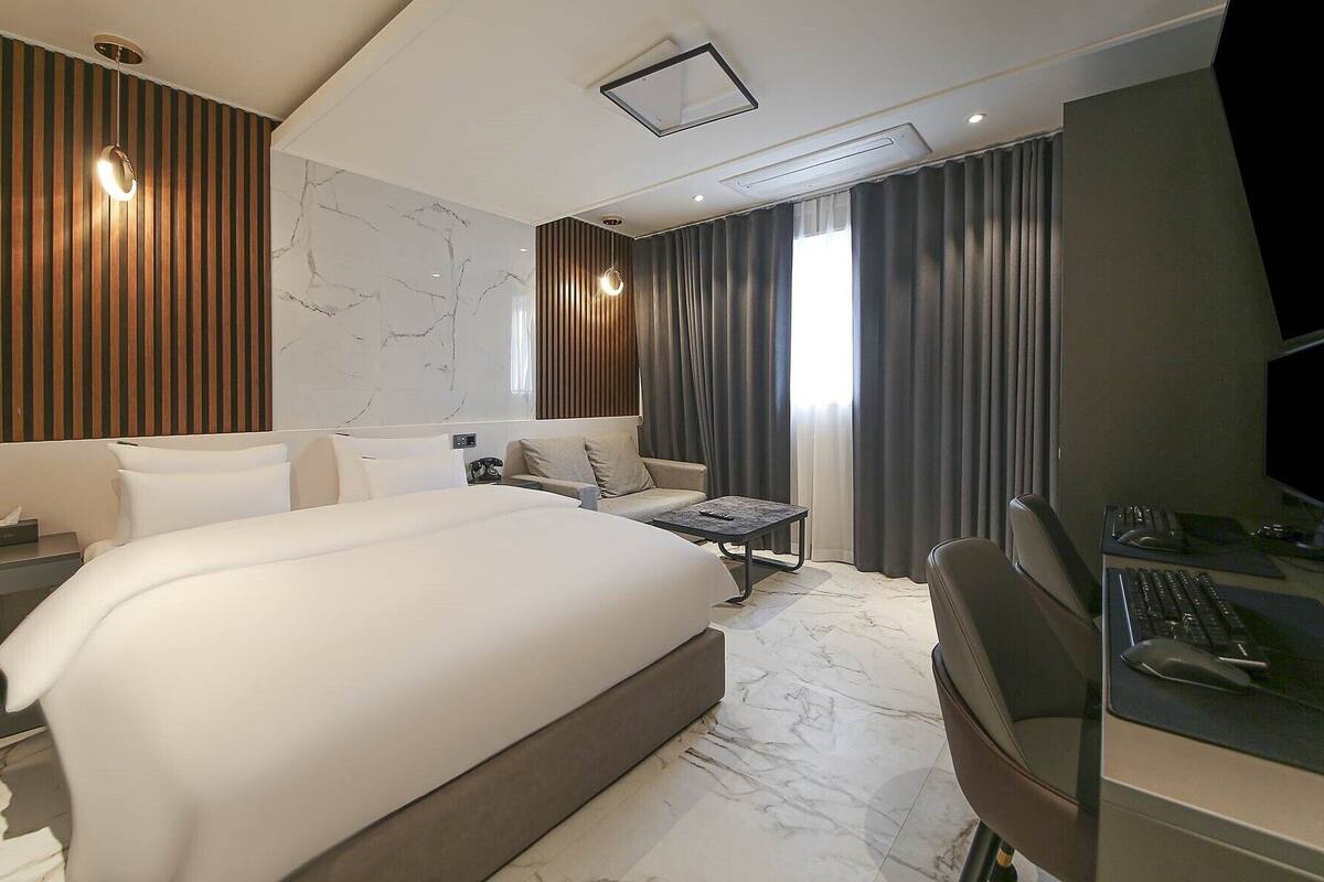 Quarto de hotel moderno com uma cama grande, uma escrivaninha com computador, uma cadeira e uma televisão.