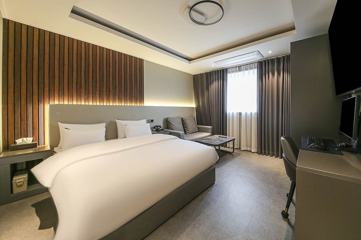 Quarto de hotel moderno com uma cama grande, uma escrivaninha com computador, televisão e janela com cortinas.