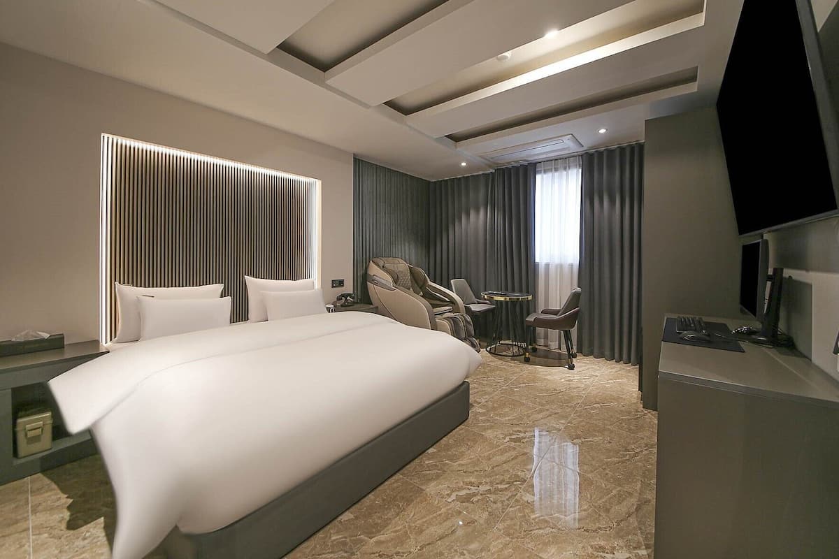 Quarto de hotel moderno com uma cama grande, uma cadeira de massagem, uma mesa com computador e uma TV.