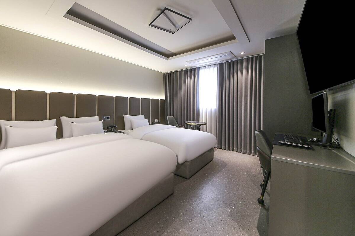 Quarto de hotel com duas camas, uma mesa de escritório com computador, televisão e janela com cortinas.