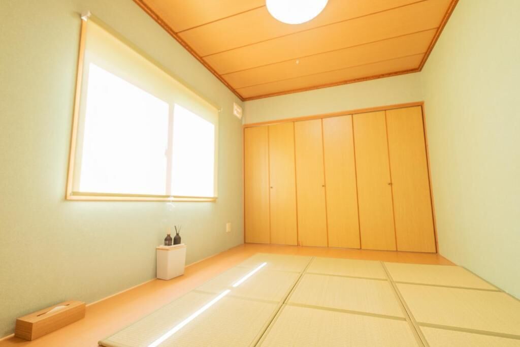 Quarto com piso de tatame, uma janela grande, um guarda-roupa de madeira e uma pequena mesa com uma garrafa.