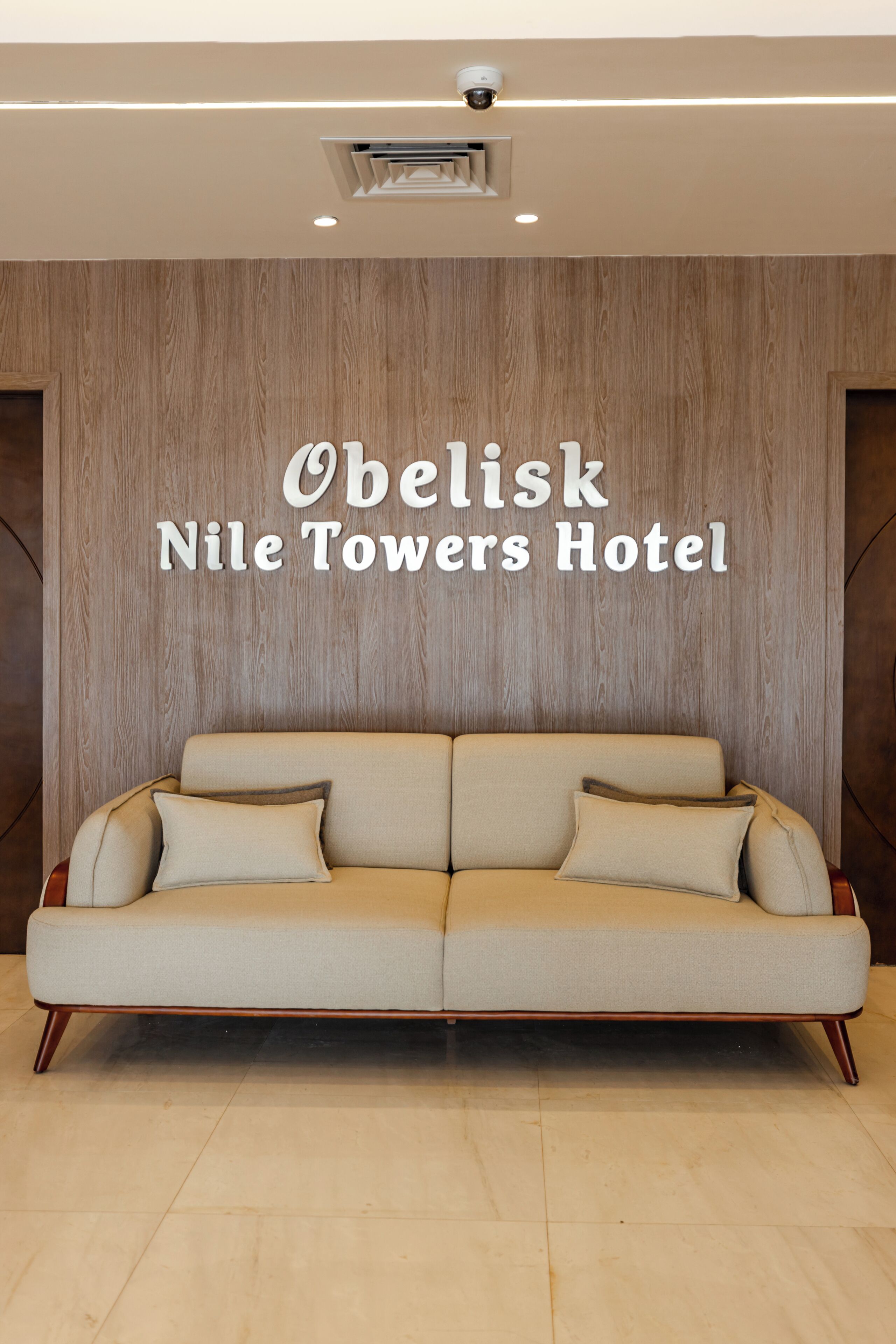 Um sofá bege num lobby de hotel com parede de madeira e uma placa que diz 'Obelisk Nile Towers Hotel'.