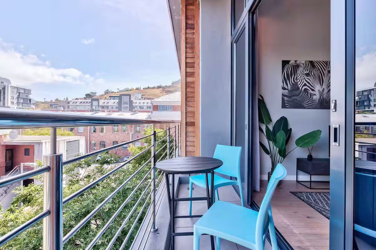 Uma varanda com vista para edifícios, uma mesa, duas cadeiras e uma planta grande.