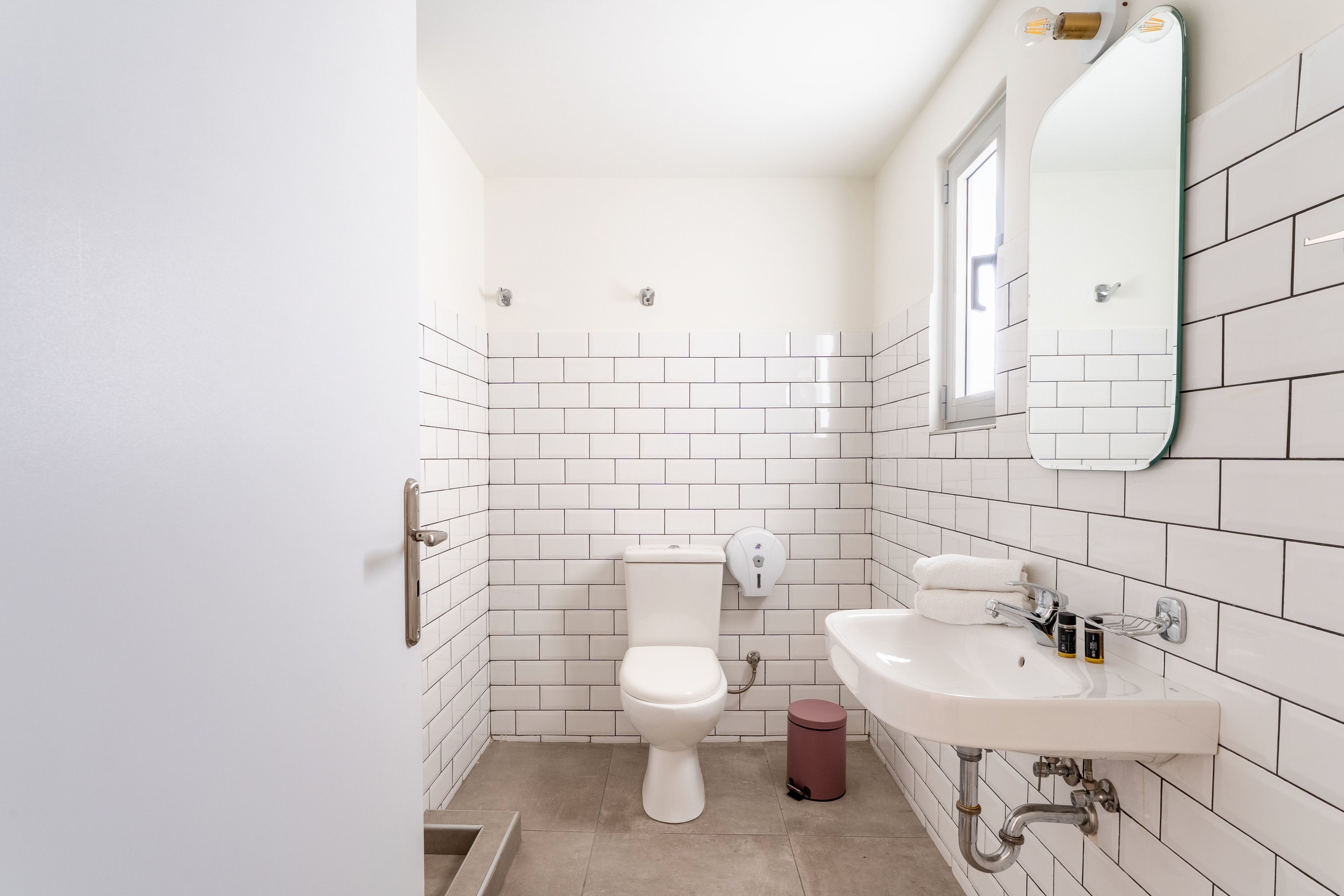 Uma casa de banho moderna com azulejos brancos, sanita, lavatório e espelho.