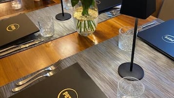 Mesa de restaurante de hotel posta com jogos americanos pretos, talheres e um vaso de rosas laranjas.