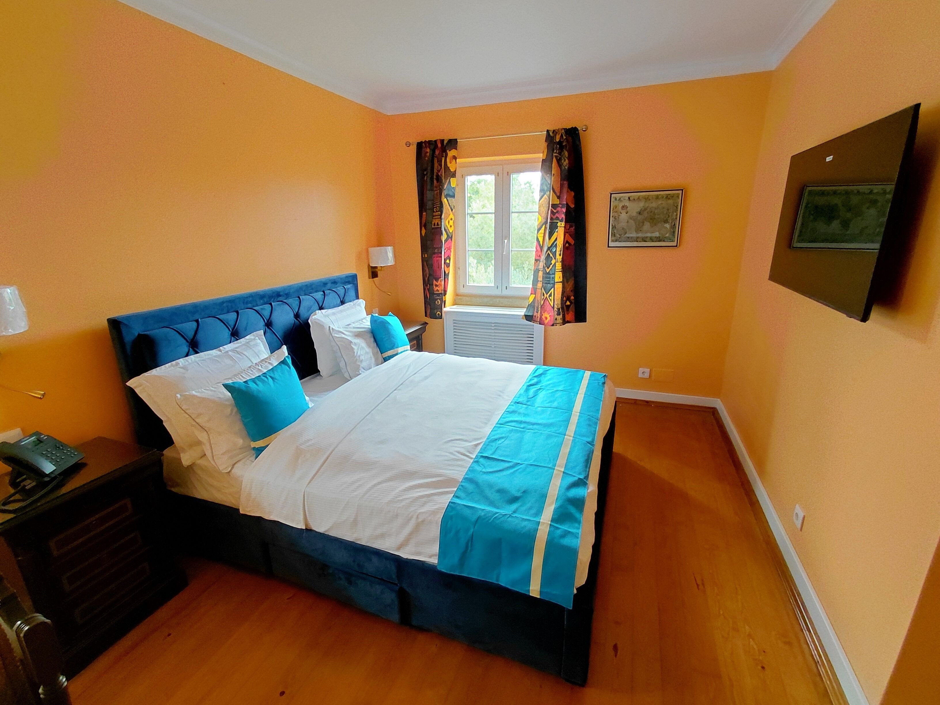 Quarto com cama grande, mesa de cabeceira, janela com cortinas e um quadro na parede.