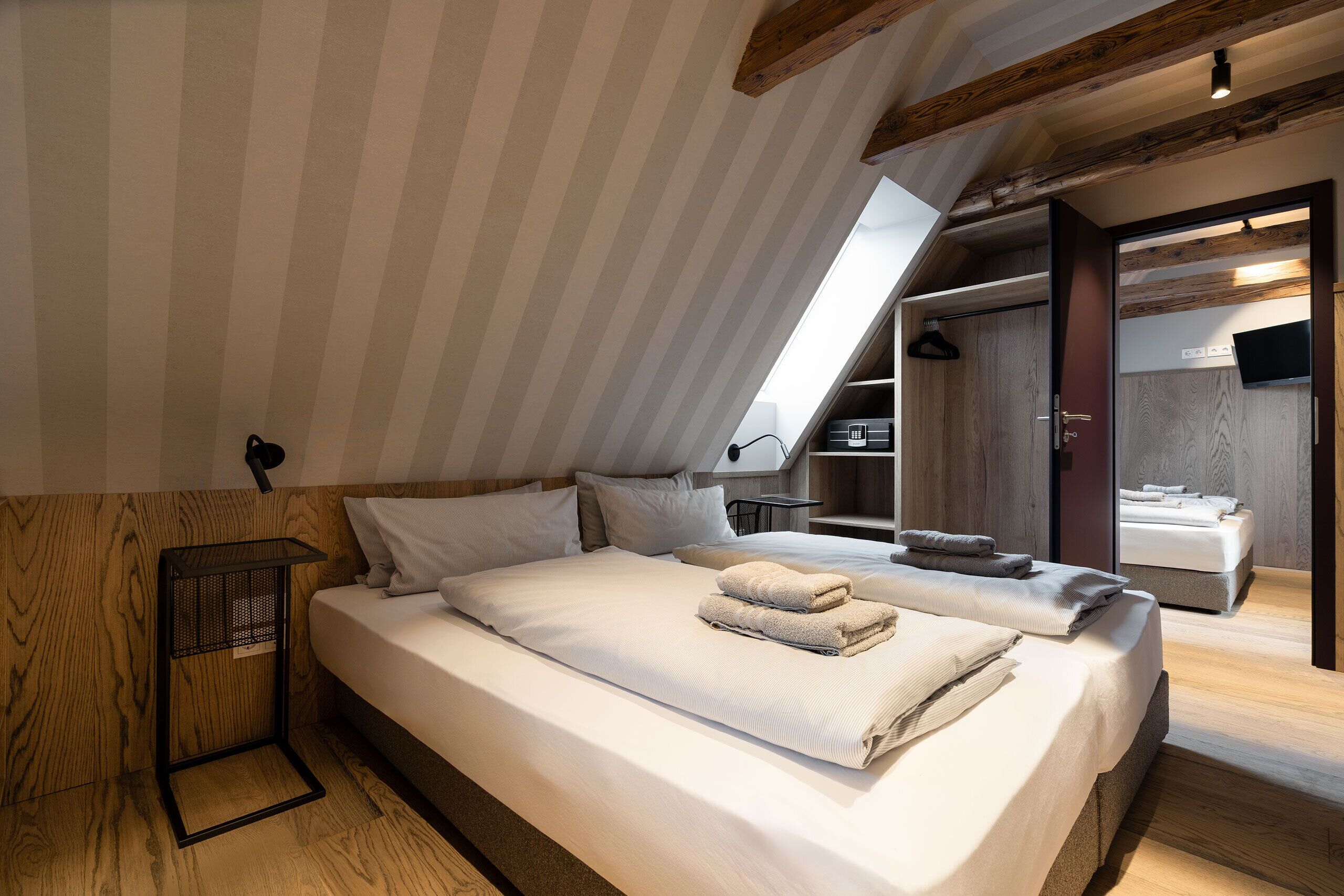 Quarto com teto inclinado, cama com roupa branca, mesa de cabeceira e piso de madeira.