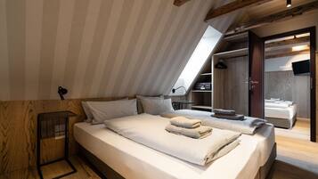 Quarto com teto inclinado, cama com roupa branca, mesa de cabeceira e piso de madeira.