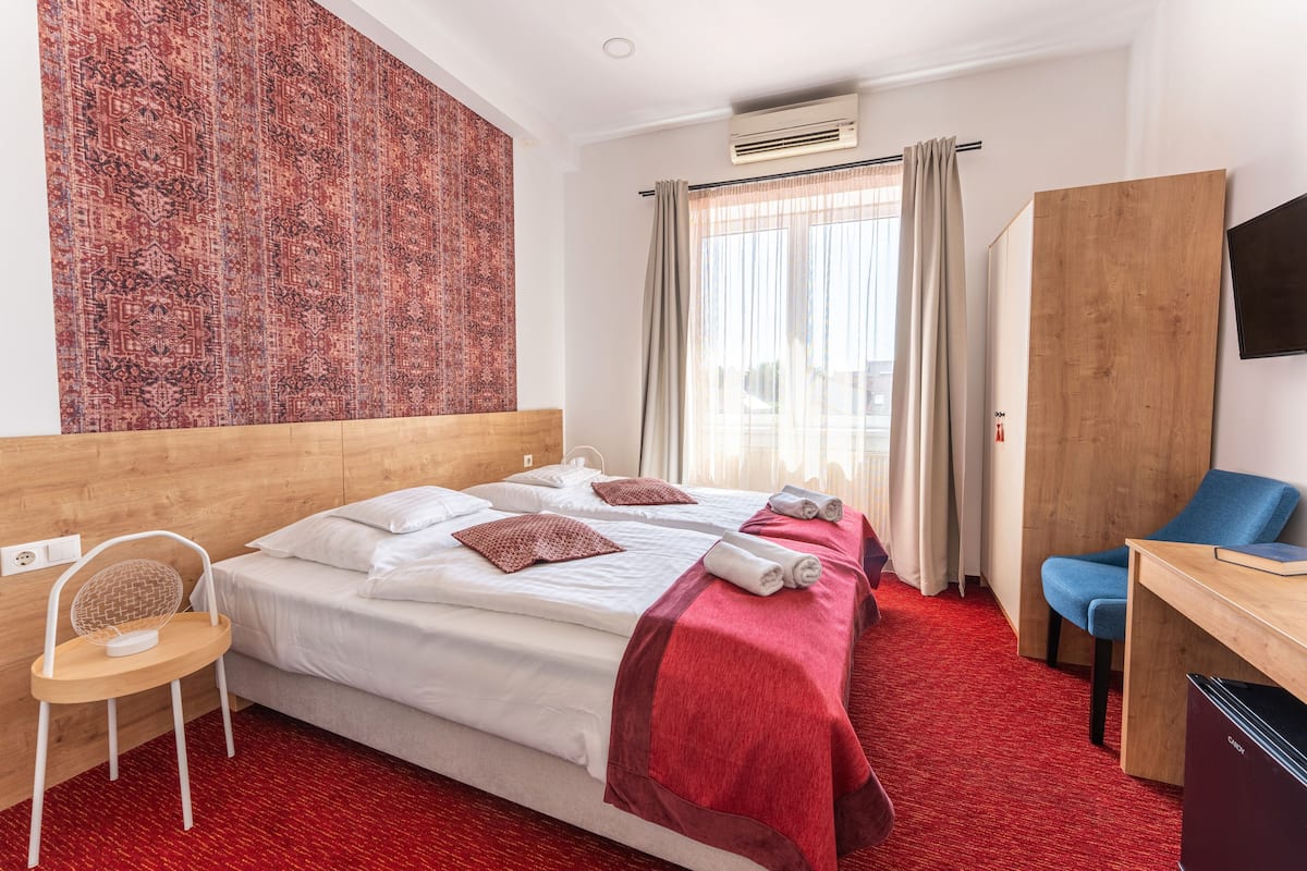 Quarto de hotel com uma cama grande, uma escrivaninha, uma cadeira, uma TV e um tapete vermelho.
