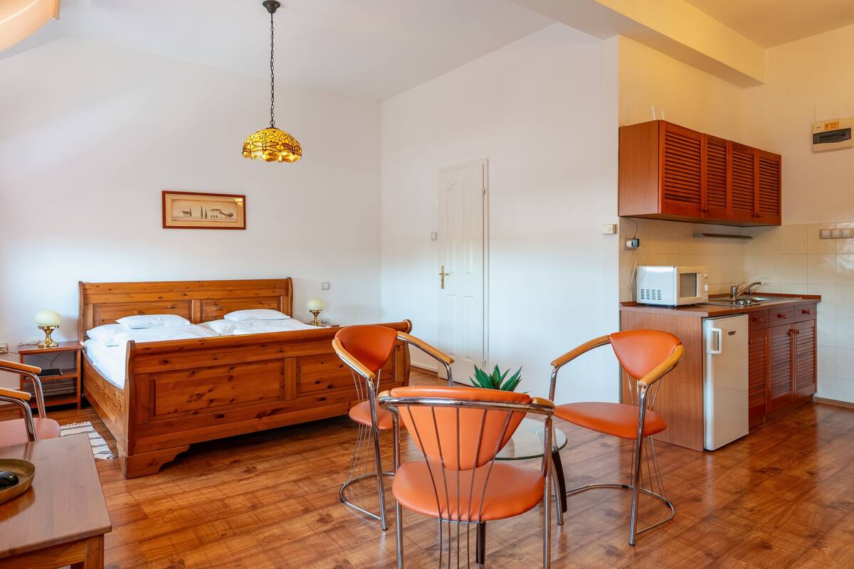 Quarto com cama de madeira, duas cadeiras laranja, uma pequena cozinha e um lustre.