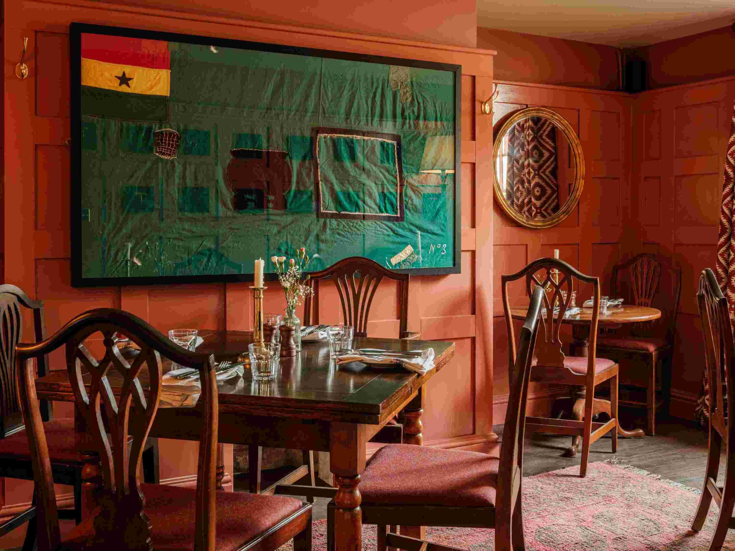 Uma sala de jantar com uma grande pintura verde, cadeiras de madeira, uma mesa posta para quatro e um tecido decorativo na parede.