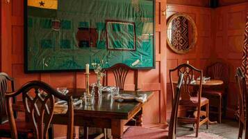 Uma sala de jantar com uma grande pintura verde, cadeiras de madeira, uma mesa posta para quatro e um tecido decorativo na parede.