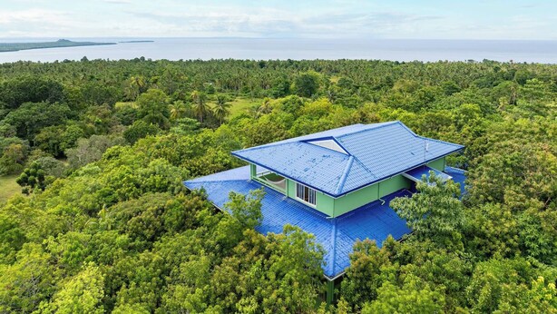 Uma casa com telhado azul cercada por uma floresta verde densa e vista para o oceano.