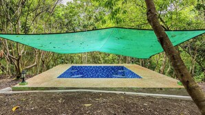 Área de piscina com chão de azulejos azuis, cercada por árvores e um dossel verde.