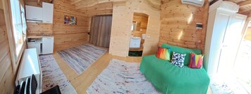 Quarto de cabana de tronco com cama verde, pequeno espaço de cozinha, banheiro e um quadro na parede.