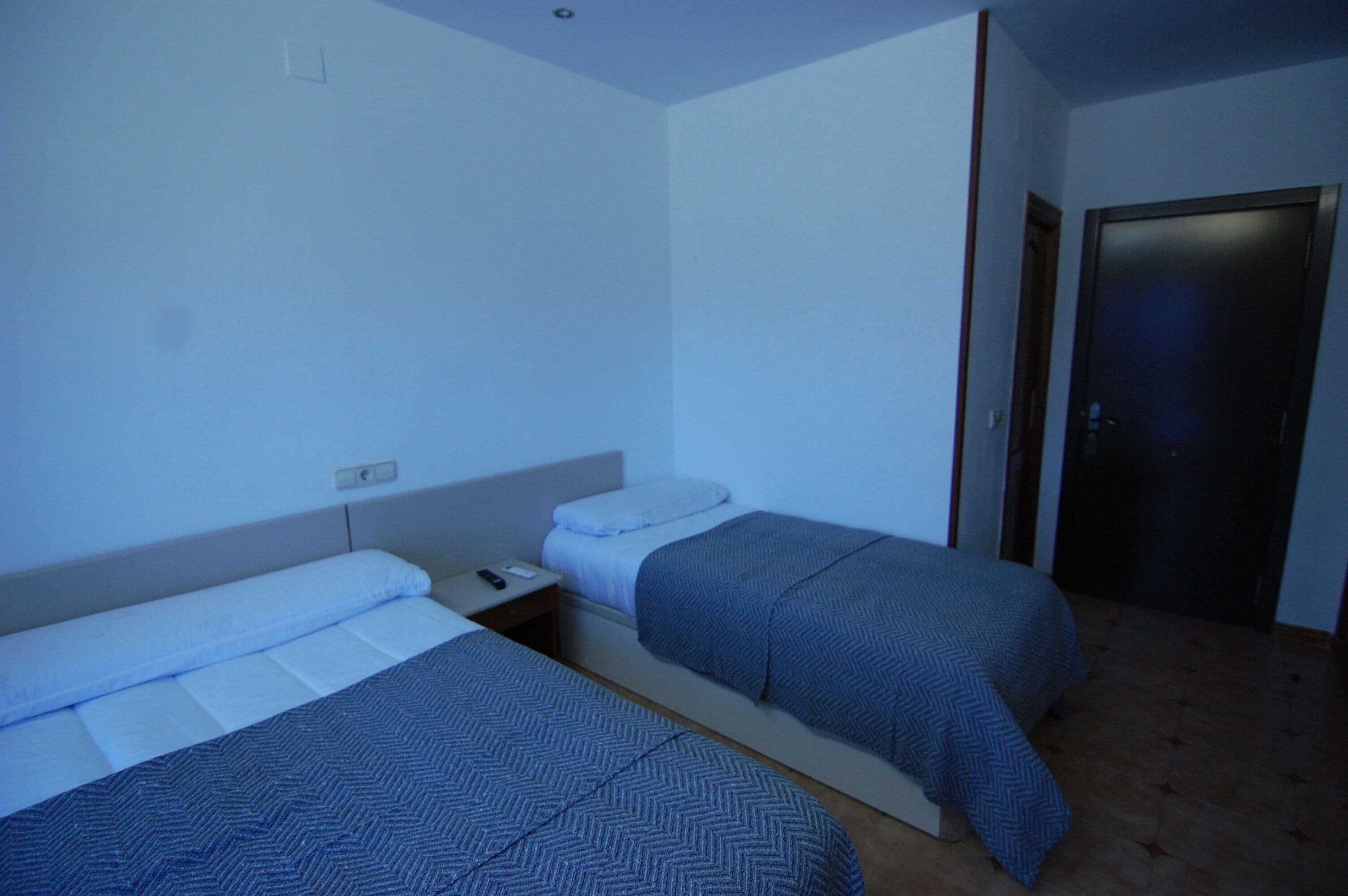 Quarto pequeno e compacto com duas camas de solteiro, uma mesinha de cabeceira, uma tomada e uma porta.
