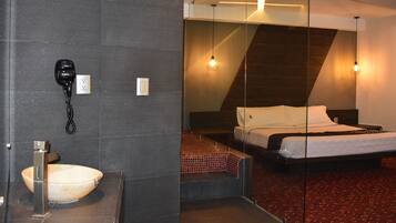 Quarto de hotel moderno com um espelho grande, uma mesa de cabeceira com uma tigela, uma cama com roupa de cama branca e um tapete vermelho.