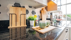 Uma cozinha com bancada de granito preto, tábua de corte de madeira, caneca verde, planta em vaso e painel de azulejos hexagonais.