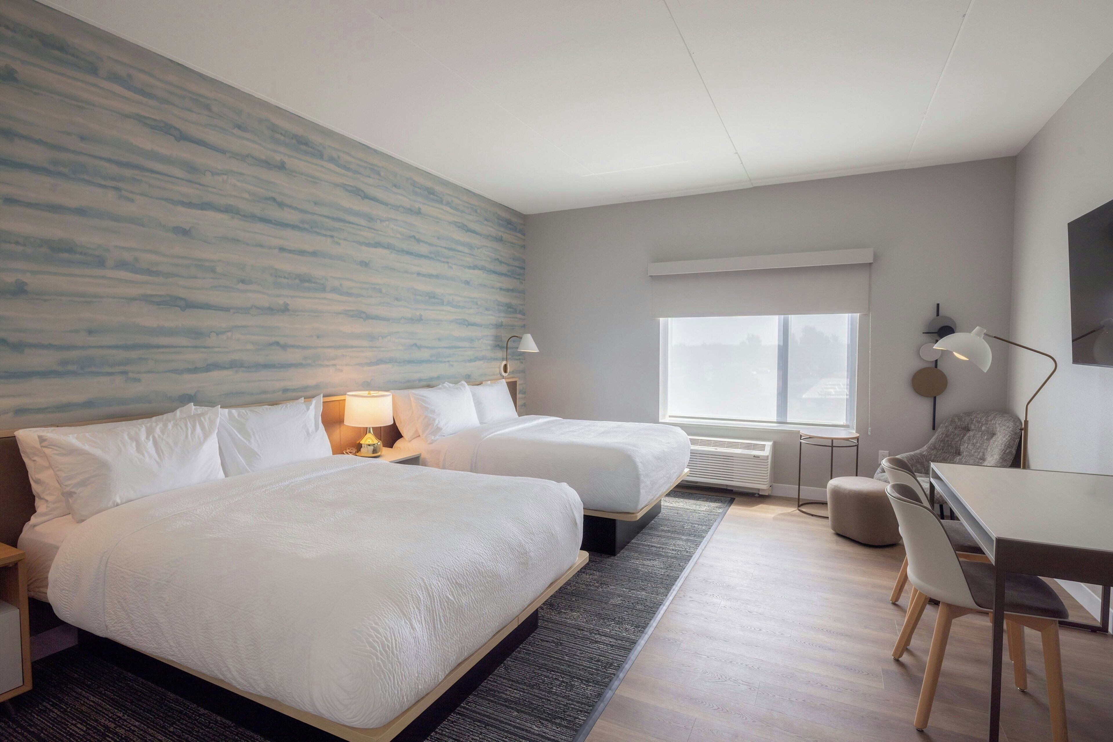 Quarto de hotel com duas camas, uma escrivaninha, uma cadeira, um abajur e uma janela grande.