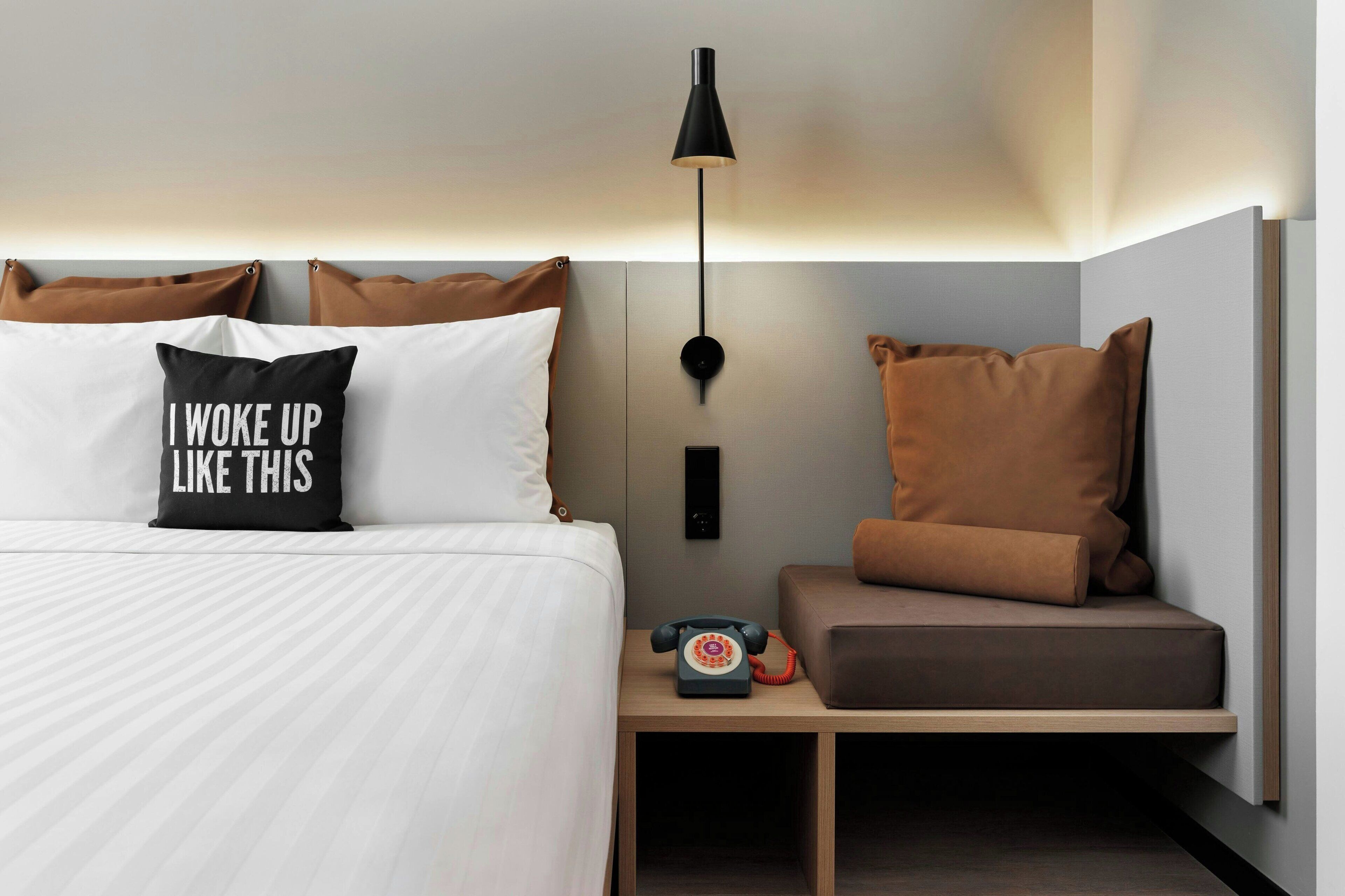 Quarto de hotel moderno com uma cama, uma mesa de cabeceira com telefone e uma luminária na parede.