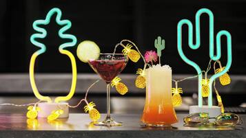 Uma área de bar com três coquetéis, cada um decorado com uma fatia de abacaxi, e luzes de neon em formato de abacaxi e cacto.