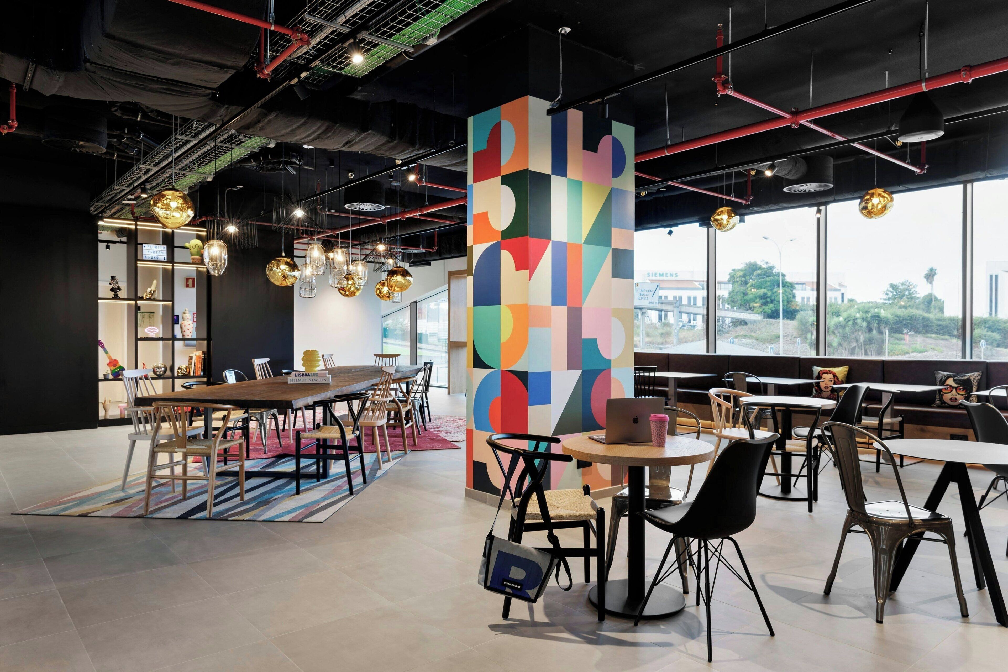 Uma sala de jantar moderna com mesas e cadeiras, uma obra de arte colorida na parede e luminárias pendentes.