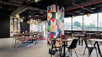 Uma sala de jantar moderna com mesas e cadeiras, uma obra de arte colorida na parede e luminárias pendentes.