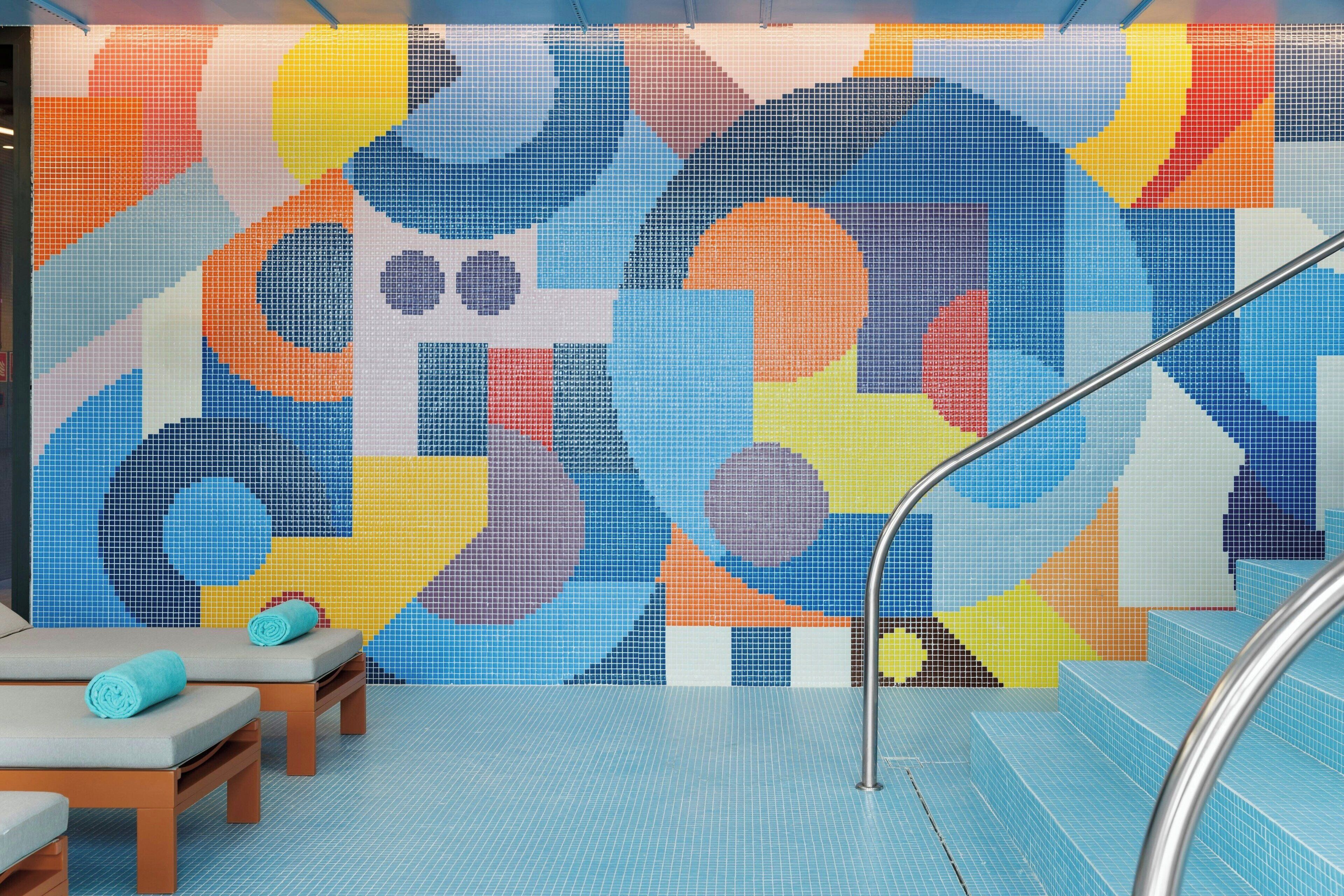 Área de piscina com parede de mosaico colorido, cadeiras de descanso e corrimão de metal.