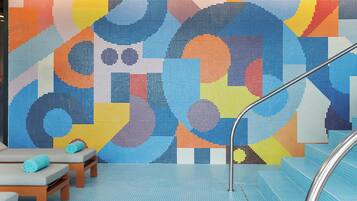 Área de piscina com parede de mosaico colorido, cadeiras de descanso e corrimão de metal.